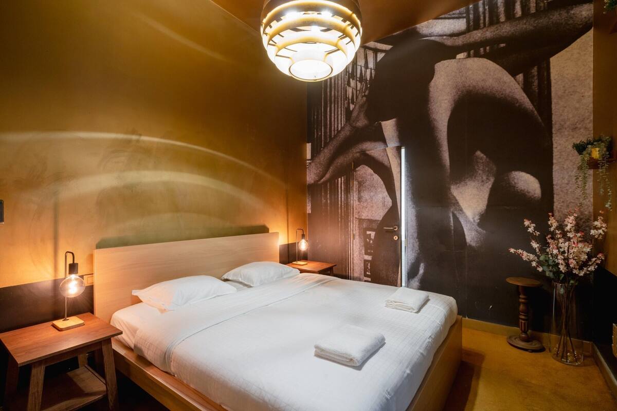 Quarto de hotel com cama, abajures, um mural na parede, uma mesinha e um vaso com flores.