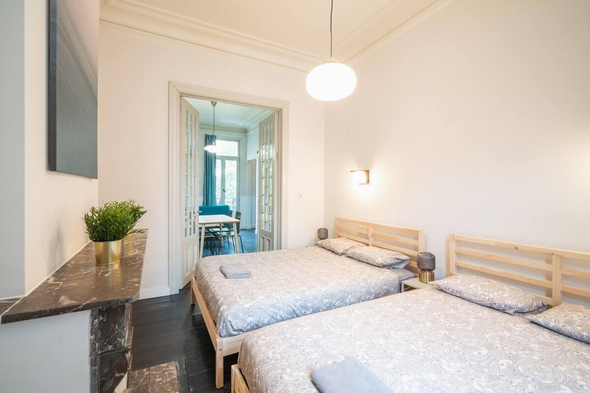 Quarto com duas camas, mesa de cabeceira, luminária, planta e vista para uma sala de jantar.