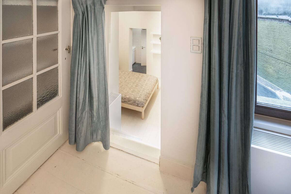 Quarto com cama, janela com cortinas e porta que dá para outro cômodo.