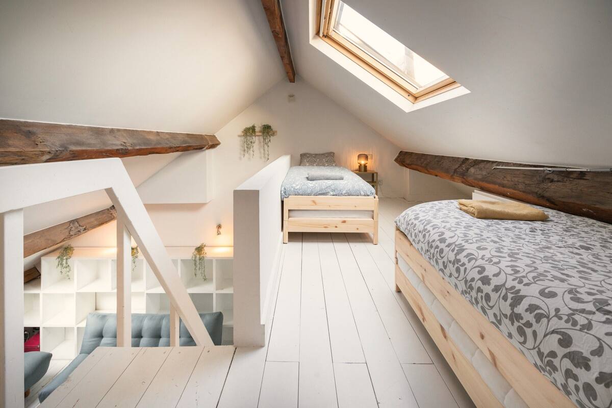 Quarto em estilo loft com claraboia, vigas de madeira, cama com roupa de cama estampada, um banco e uma pequena área de estar.