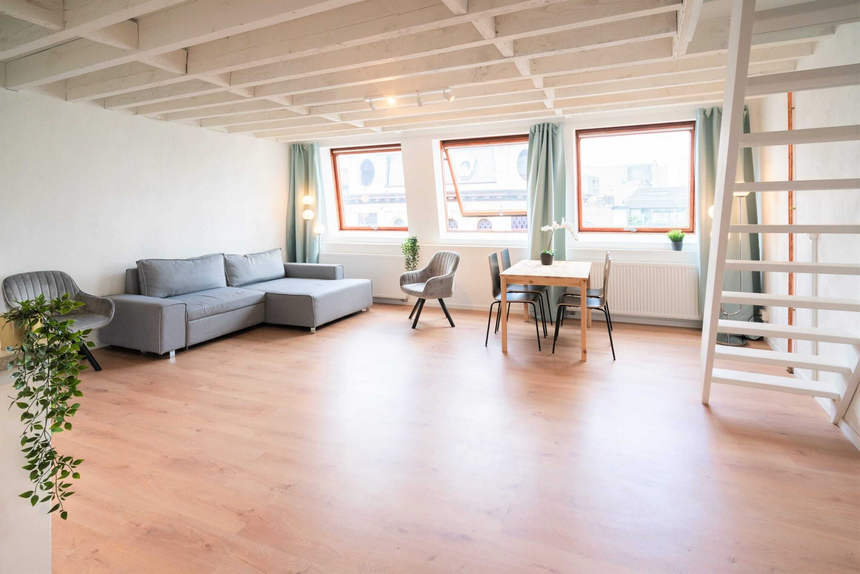 Uma sala de estar moderna com um sofá seccional cinza, uma mesa de jantar com cadeiras, uma escada branca e grandes janelas com cortinas.
