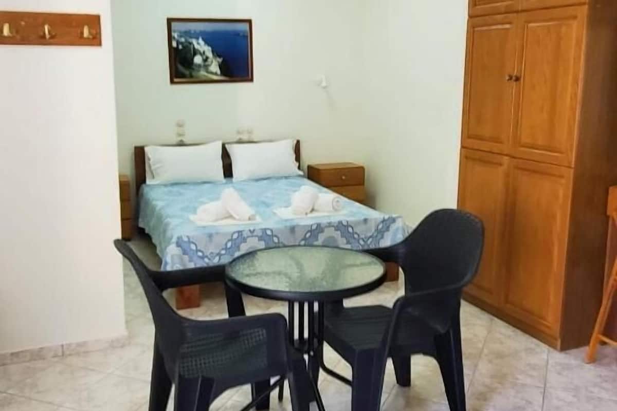Um quarto com uma cama, uma mesinha com cadeiras, um guarda-roupa de madeira e um quadro na parede.
