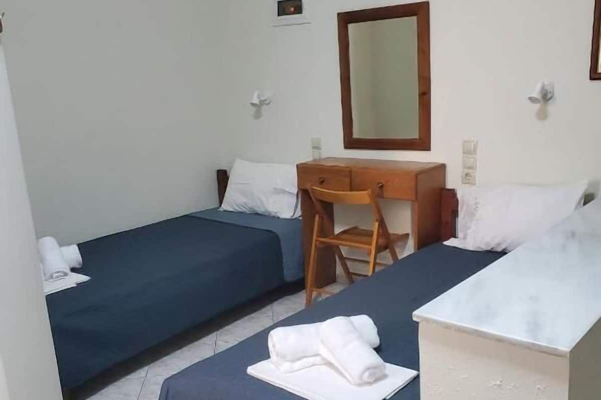 Quarto pequeno e limpo com duas camas de solteiro, uma escrivaninha de madeira, um espelho e uma pequena luz na parede.