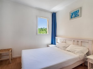 Um quarto com cama, mesa de cabeceira, janela com vista para o verde e um quadro na parede.