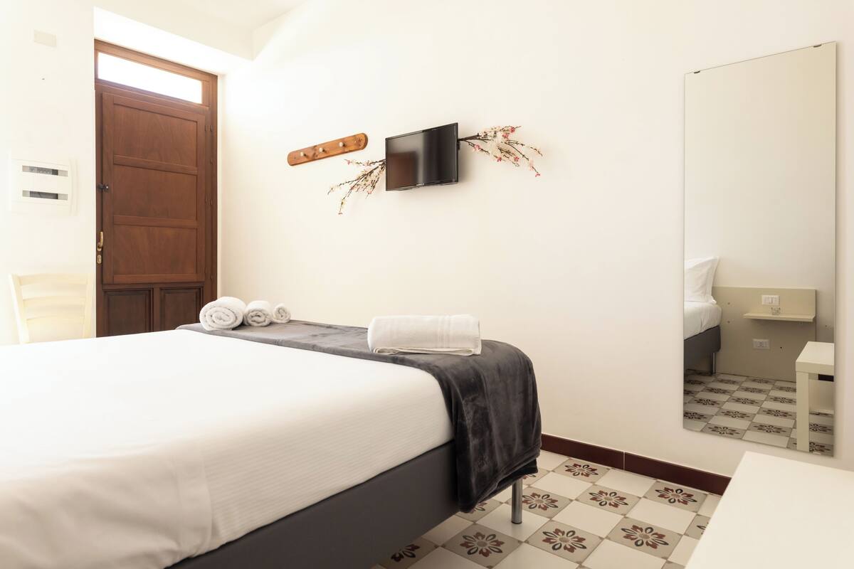 Quarto de hotel com cama, televisão na parede, porta de madeira e espelho de banheiro.