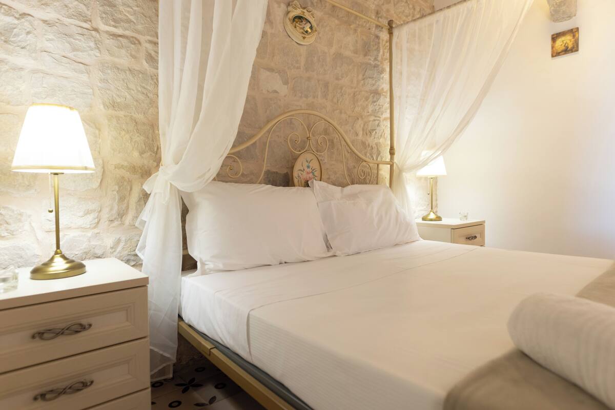 Uma cama com lençóis brancos, uma mesa de cabeceira com abajur e uma parede de pedra.