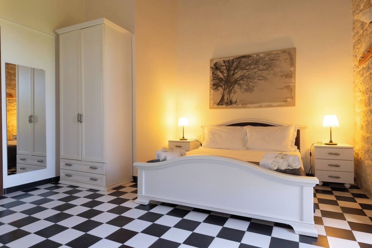 Quarto com piso xadrez, uma cama branca, duas mesas de cabeceira com abajures, um guarda-roupa e um quadro na parede.