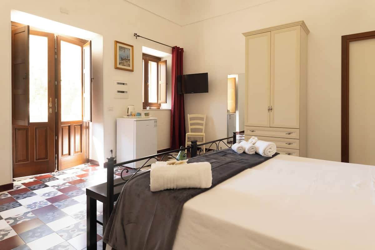 Quarto de hotel com cama, escrivaninha, cadeira, guarda-roupa, televisão e banheiro visível por uma porta aberta.
