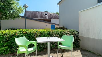 Um pátio com duas cadeiras verdes e uma mesa pequena, uma placa de identificação e um edifício com telhado inclinado ao fundo.