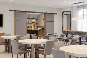 Uma sala de jantar moderna com mesas e cadeiras redondas, cozinha ao fundo e um espelho grande com iluminação decorativa.