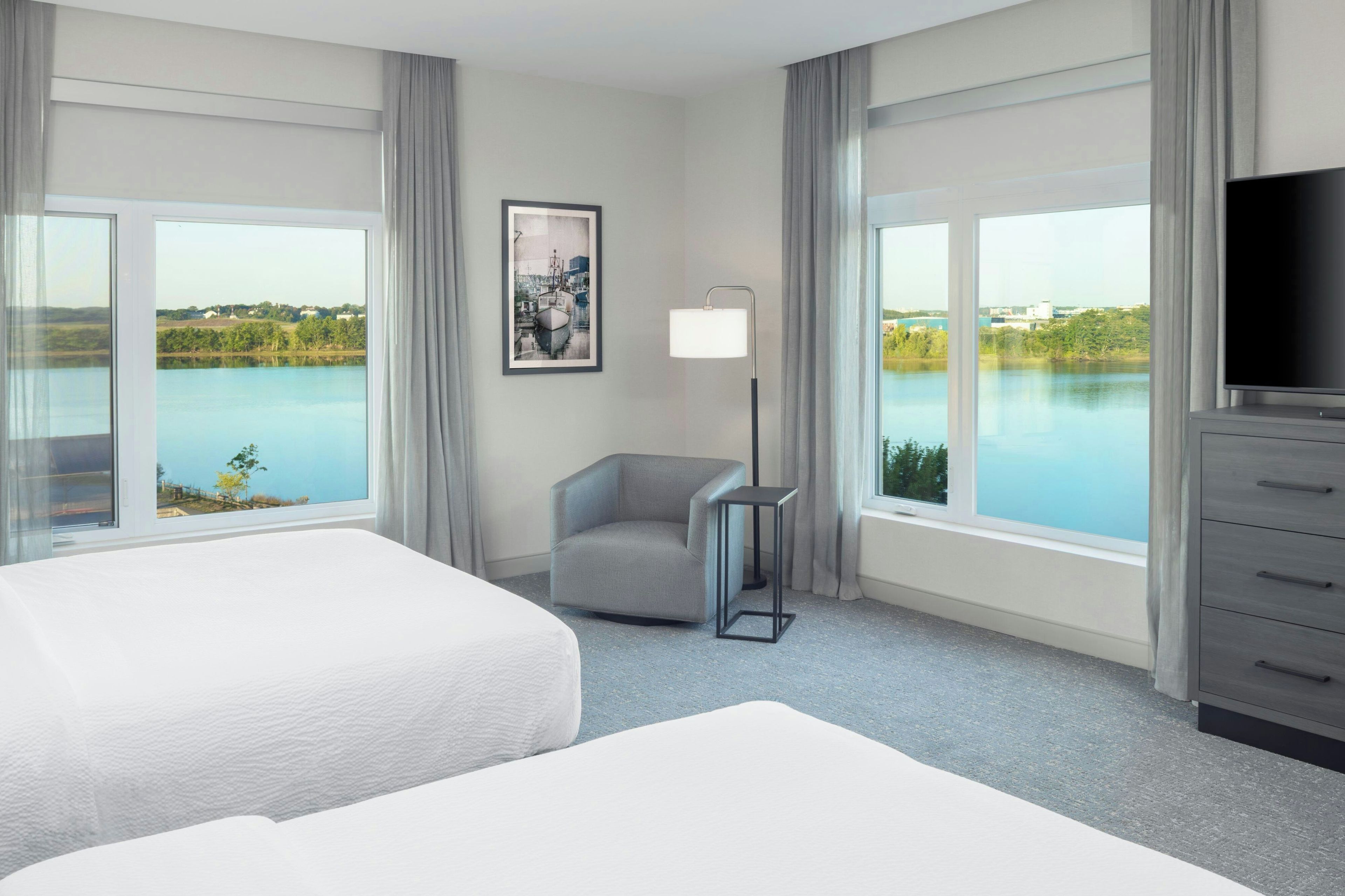 Quarto de hotel com duas camas, uma escrivaninha, uma cadeira, televisão e janelas amplas com vista para a água.