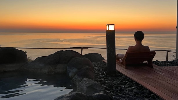 Uma pessoa sentada em um deck de madeira apreciando a vista para a água ao pôr do sol.