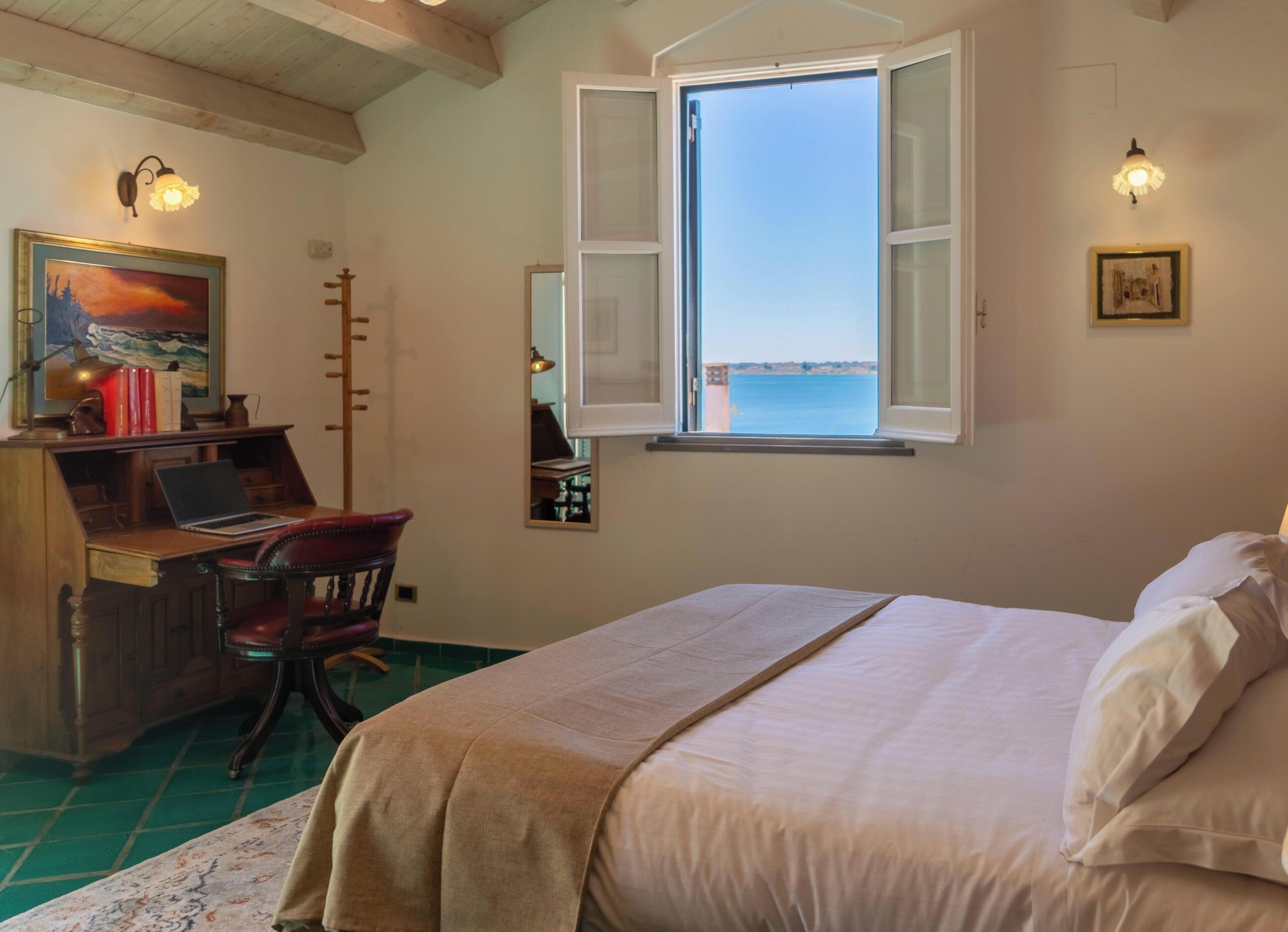 Quarto com cama, escrivaninha com laptop, cadeira, janela com vista e pintura na parede.