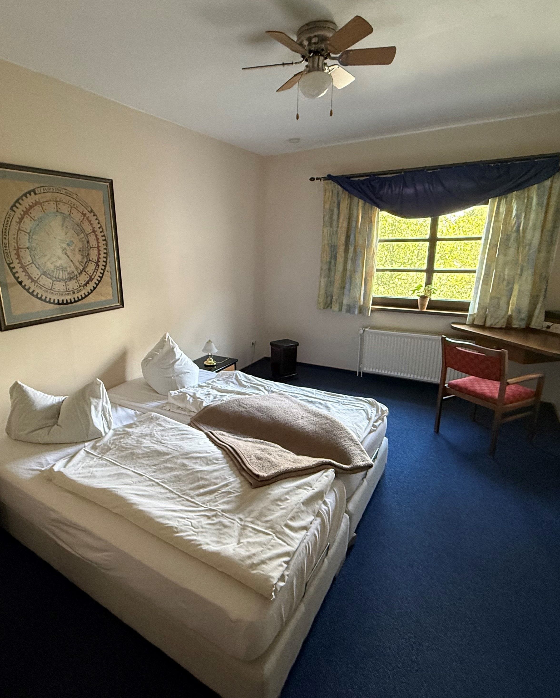 Quarto com cama, escrivaninha, cadeira, janela com cortinas, radiador e ventilador de teto.