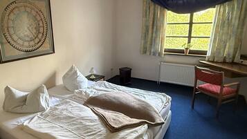 Quarto com cama, escrivaninha, cadeira, janela com cortinas, radiador e ventilador de teto.