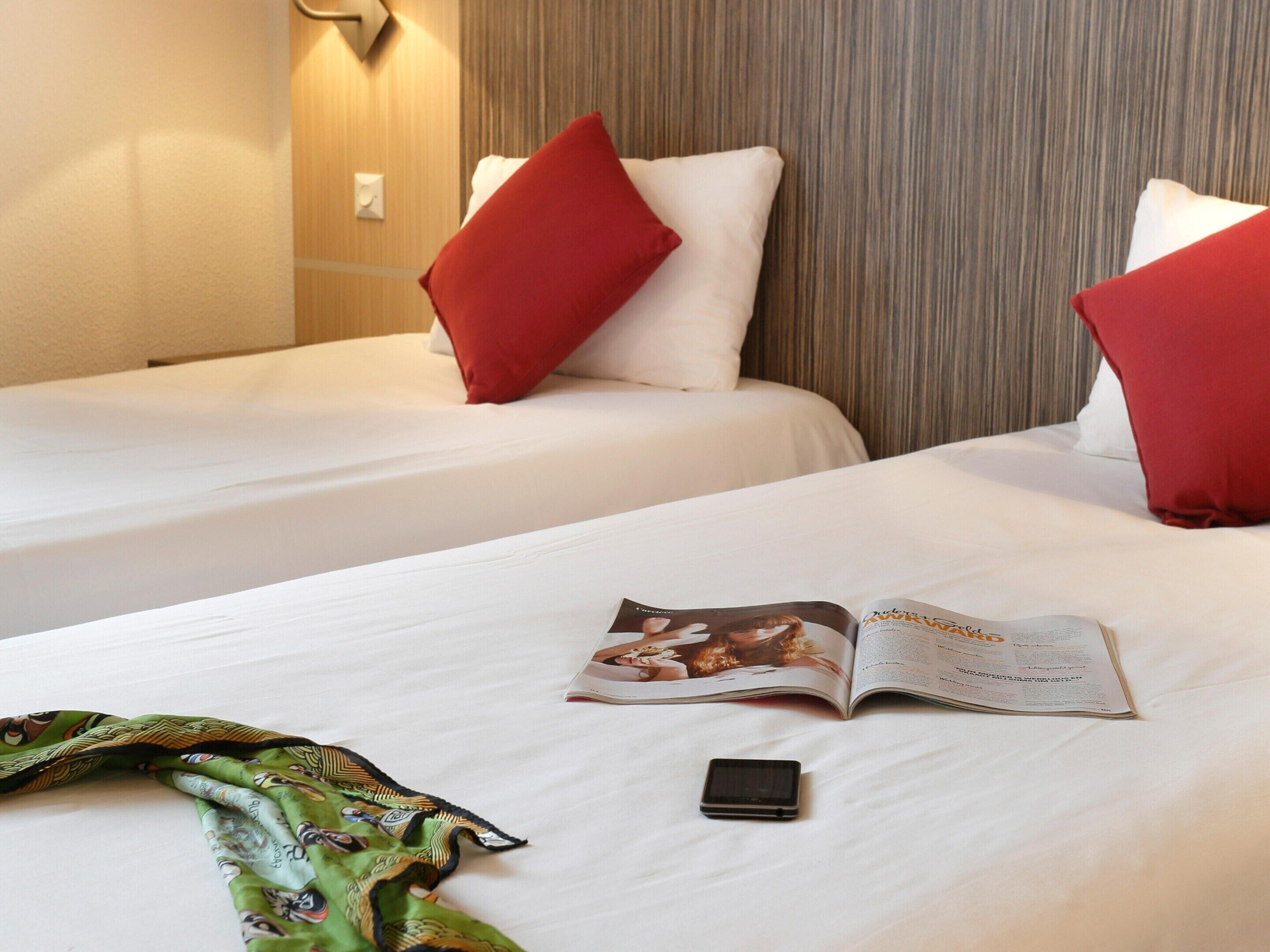 Quarto de hotel com duas camas, cada uma com roupa de cama branca e almofadas vermelhas, uma revista e um smartphone na cama.