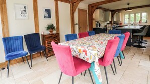 Uma sala de jantar com uma mesa comprida, cadeiras coloridas, vigas de madeira e uma cozinha ao fundo.