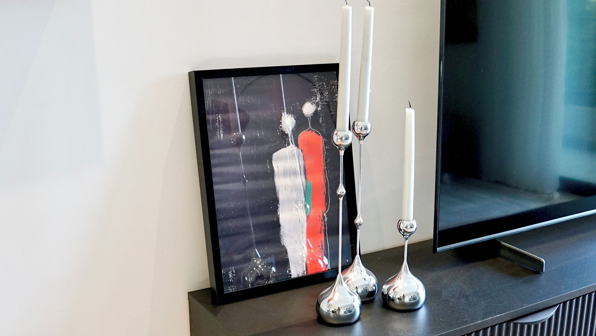 Uma obra de arte emoldurada com figuras abstratas, dois castiçais metálicos e uma janela moderna.