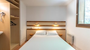 Quarto de hotel pequeno e estreito, com uma cama de solteiro, mesas de cabeceira, um guarda-roupa e uma janela.
