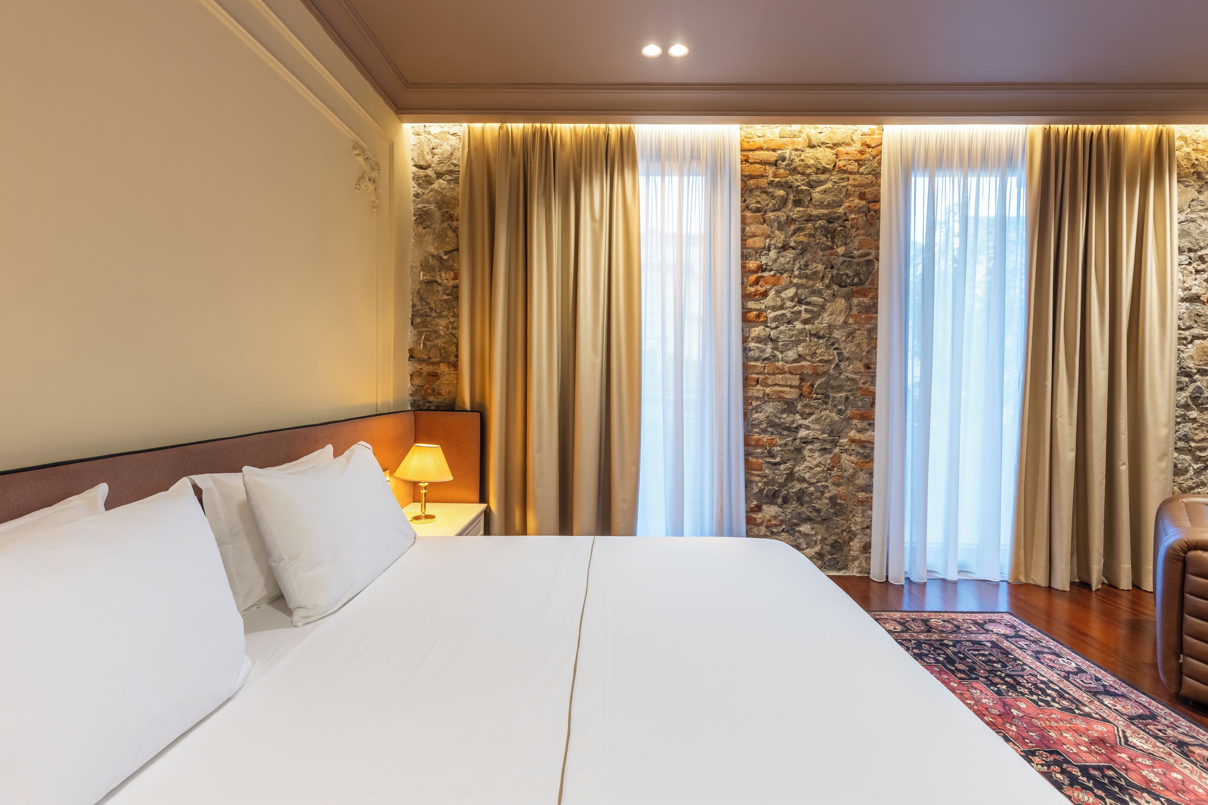 Quarto de hotel com uma cama grande, mesa de cabeceira com abajur, parede de pedra e vista através de cortinas translúcidas.
