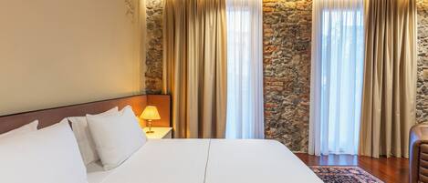 Quarto de hotel com uma cama grande, mesa de cabeceira com abajur, parede de pedra e vista através de cortinas translúcidas.