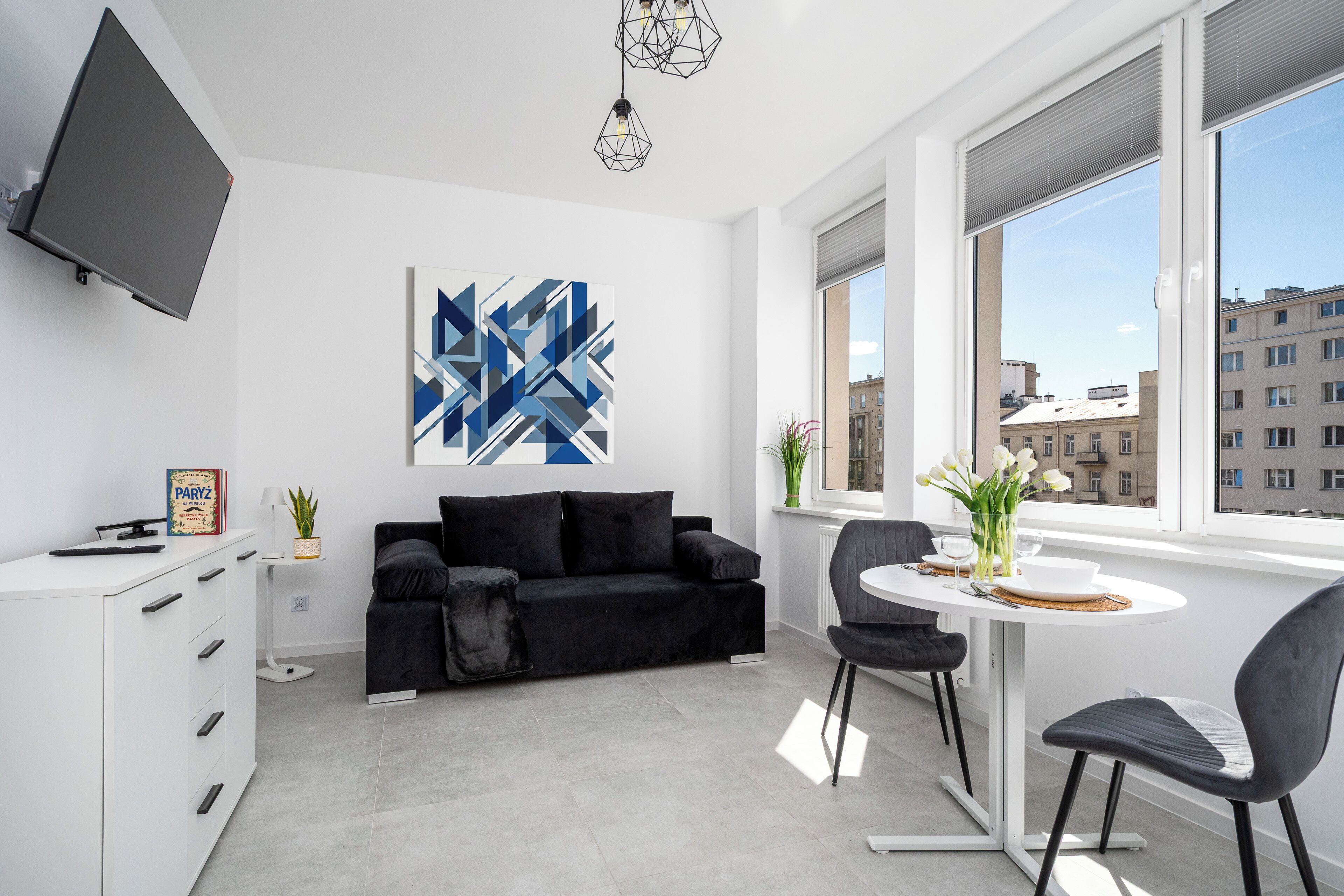 Uma sala de estar moderna com um sofá preto, uma mesa branca com cadeiras, uma televisão montada na parede e um quadro abstrato grande.