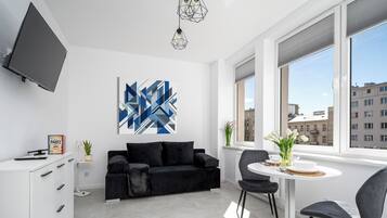 Uma sala de estar moderna com um sofá preto, uma mesa branca com cadeiras, uma televisão montada na parede e um quadro abstrato grande.