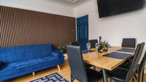 Uma sala de estar moderna com um sofá azul, uma mesa de jantar de madeira com quatro cadeiras, uma TV fixada na parede e uma porta com moldura azul.