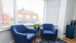 Quarto com duas poltronas azuis, um tapete estampado, uma mesa pequena com uma planta e vista para um edifício de tijolos através de grandes janelas.