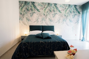 Quarto com cama, mesas de cabeceira, uma tigela de frutas e papel de parede com folhagens tropicais.