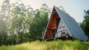 Uma cabana moderna, de madeira triangular e telhado de metal, rodeada por árvores altas e vegetação gramínea.