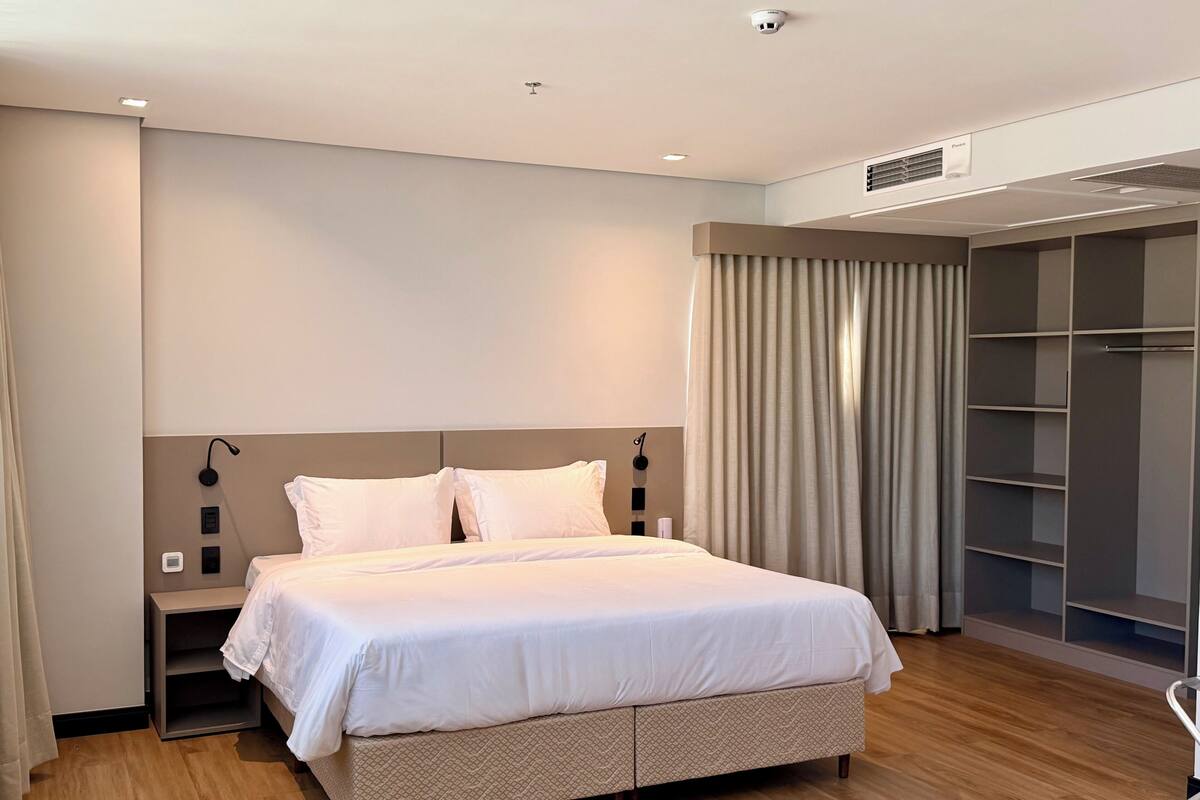 Quarto de hotel moderno com cama grande, mesas de cabeceira, guarda-roupa e câmera de segurança instalada no teto.