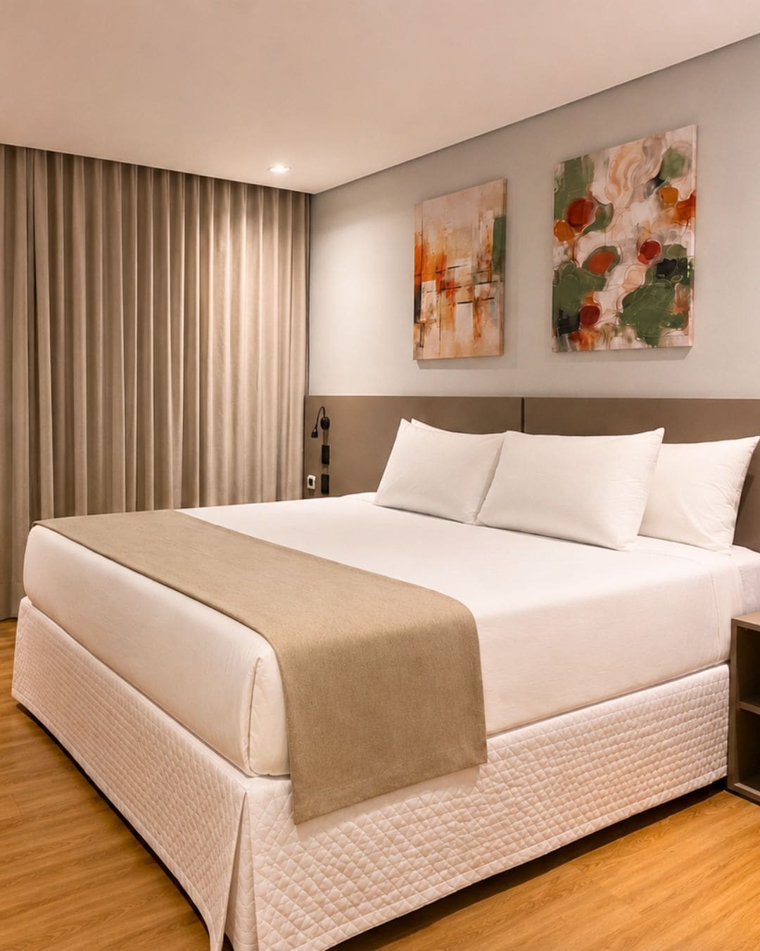 Quarto de hotel com uma cama grande, uma mesinha de cabeceira e duas pinturas abstratas na parede.