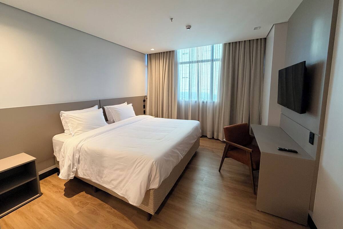 Quarto de hotel com uma cama grande, uma escrivaninha, uma cadeira, uma televisão e uma janela com cortinas.