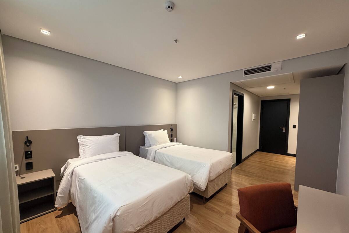 Quarto de hotel com duas camas, uma escrivaninha, uma cadeira e a porta do banheiro.