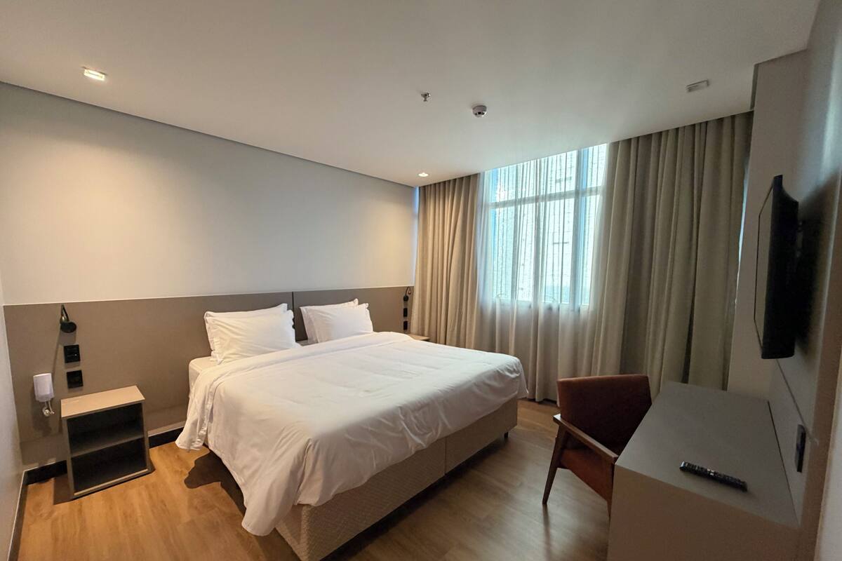 Quarto de hotel com cama, escrivaninha, cadeira, televisão e janela com cortinas.