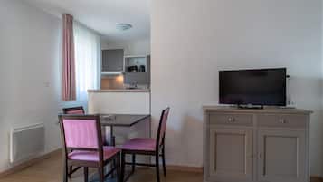 Quarto compacto com uma mesa de jantar, duas cadeiras, uma televisão sobre um móvel e uma pequena cozinha ao fundo.