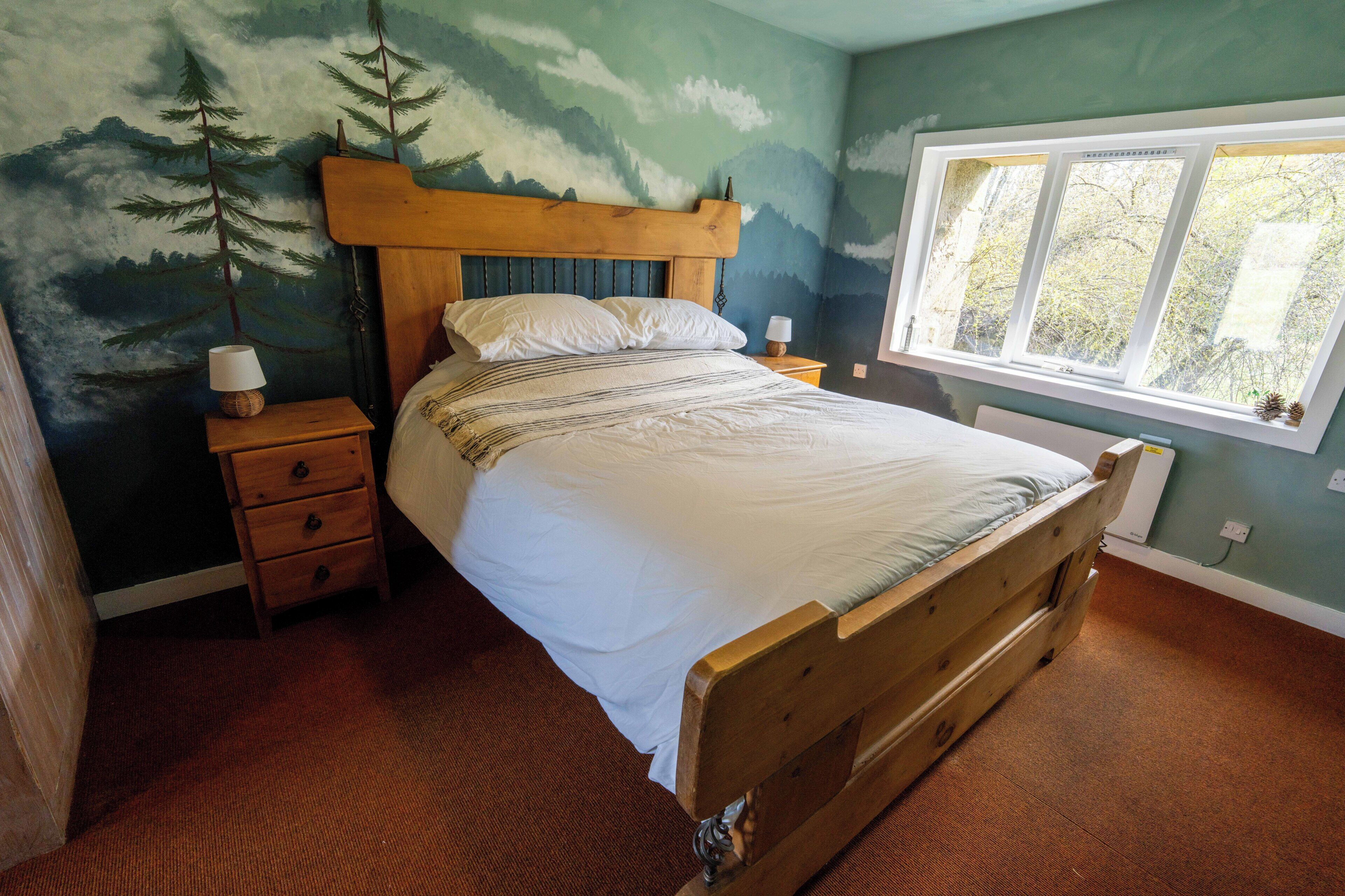 Quarto com cama de madeira, mesa de cabeceira com abajur, cômoda e janela com vista para árvores.