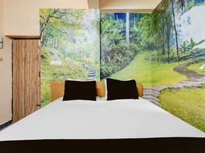 Uma cama com roupa de cama branca e almofadas pretas, um guarda-roupa de madeira e uma grande tapeçaria com tema de floresta na parede.
