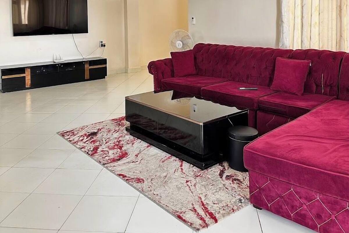 Uma sala de estar moderna com um sofá seccional, uma televisão de tela plana, um lustre e uma janela grande com cortinas.