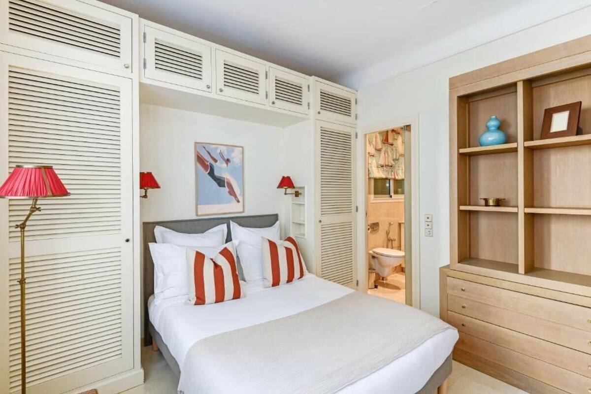 Quarto com cama, luminárias de cabeceira, um quadro, um armário de madeira e um banheiro visível por uma porta aberta.