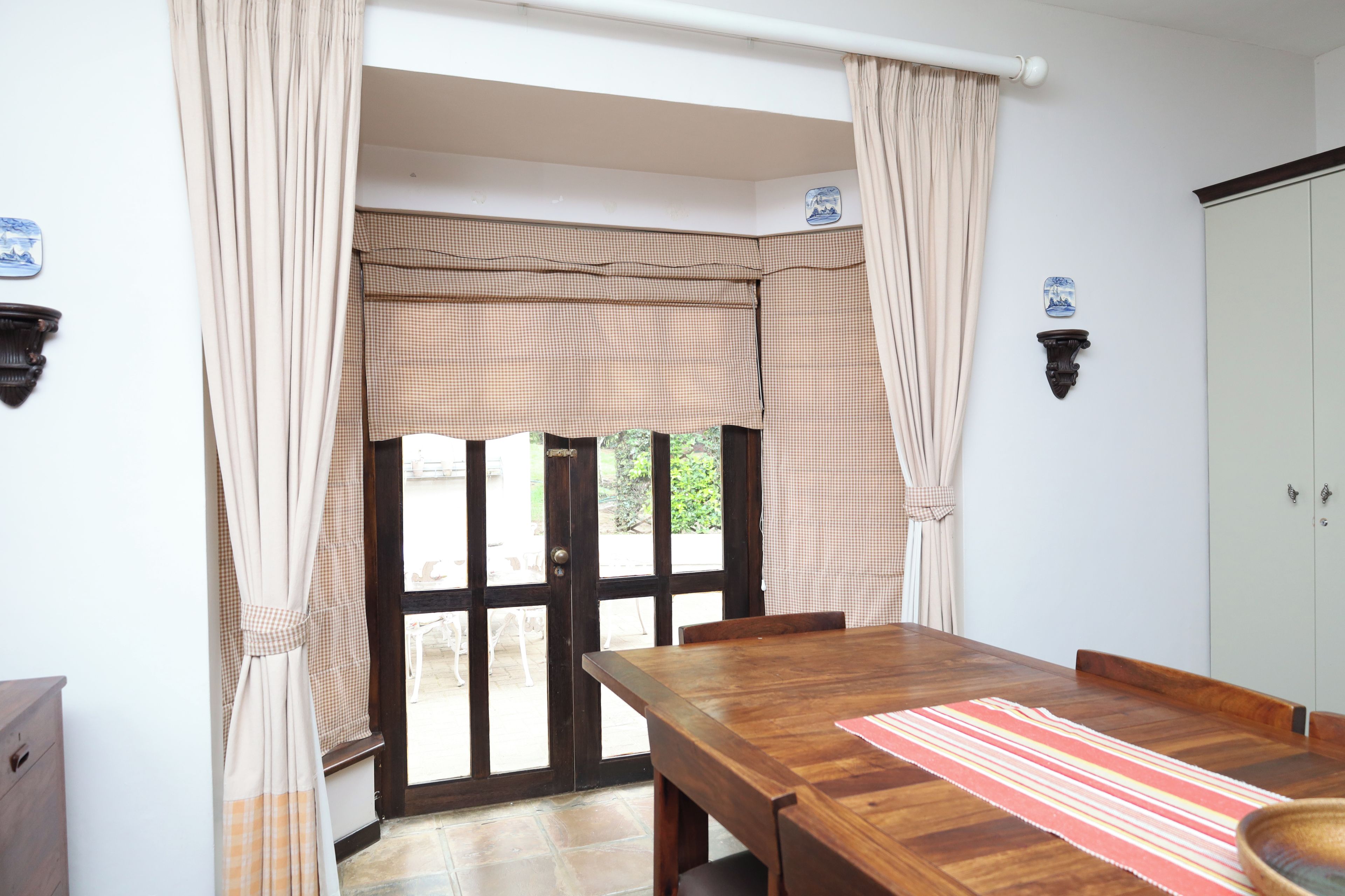 Uma sala de jantar com mesa de madeira, cortinas bege e vista por uma porta de vidro.