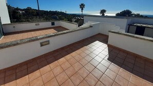 Terraço no telhado com piso de azulejos, paredes brancas e vista desobstruída para o mar e colinas.