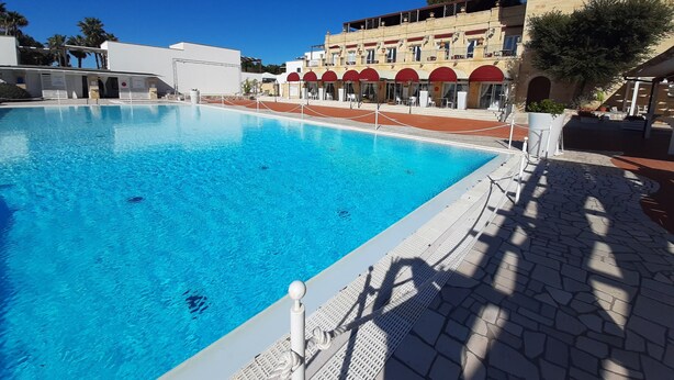 Uma piscina com superfície de água azul cristalina, rodeada por um calçadão pavimentado e um edifício com toldos vermelhos.