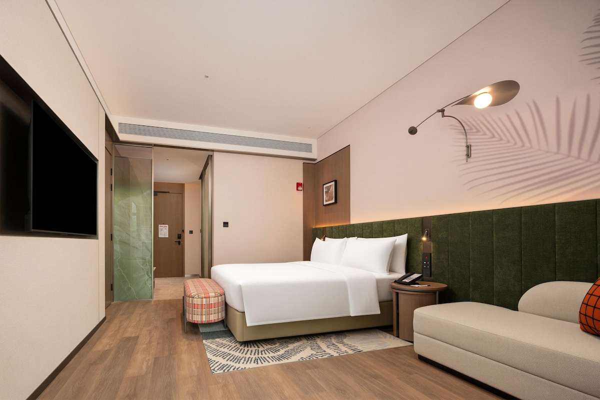 Quarto de hotel com cama, mesa de cabeceira, abajur, sofá e um banquinho.