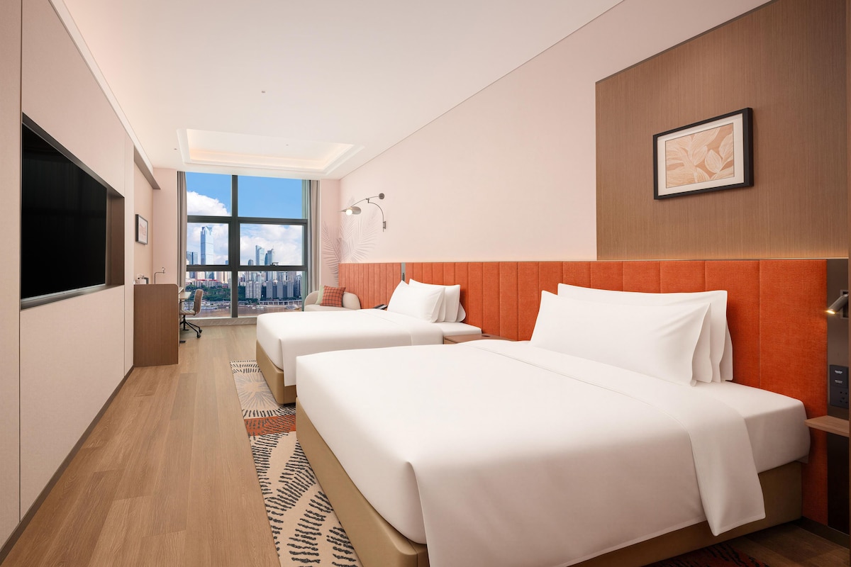 Quarto de hotel com duas camas, uma janela grande com vista para a cidade, televisão de tela plana e luminária fixada na parede.