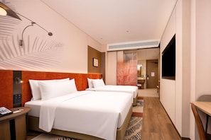Quarto de hotel com duas camas, uma mesa de trabalho, uma cadeira, uma televisão e banheiro visível através de um painel de vidro.