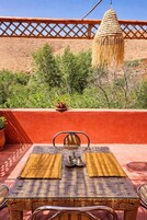 Mesa de vime com duas cadeiras, luminária pendente e plantas em vasos numa varanda com vista para arbustos verdes e uma parede vermelha.