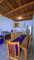 Uma mesa de jantar de madeira com cadeiras, uma área de cozinha com armários e eletrodomésticos, e um chão ladrilhado.