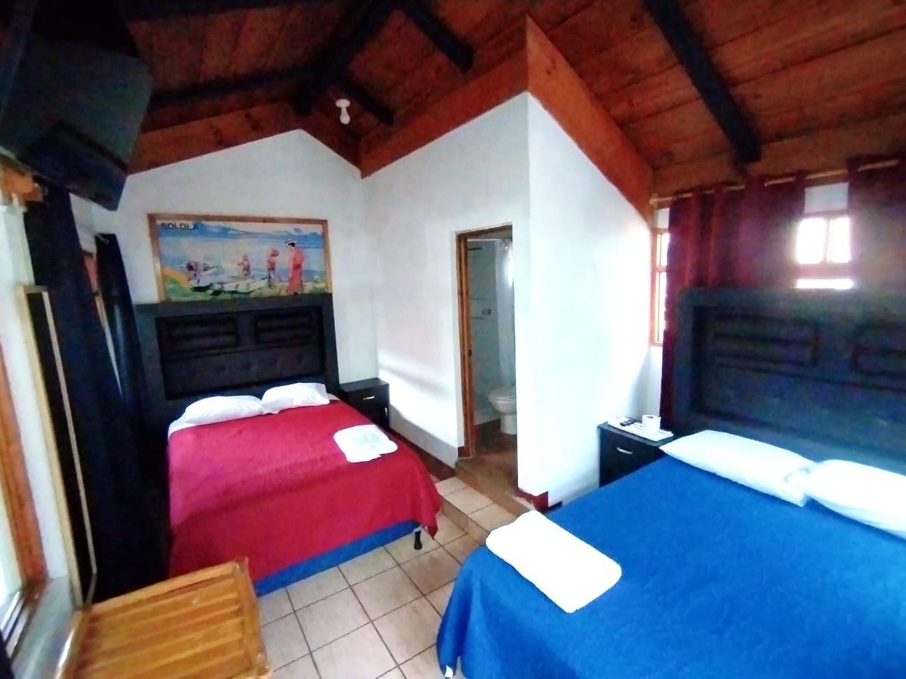 Quarto com duas camas, vigas de madeira no teto, um quadro na parede e porta para um banheiro.